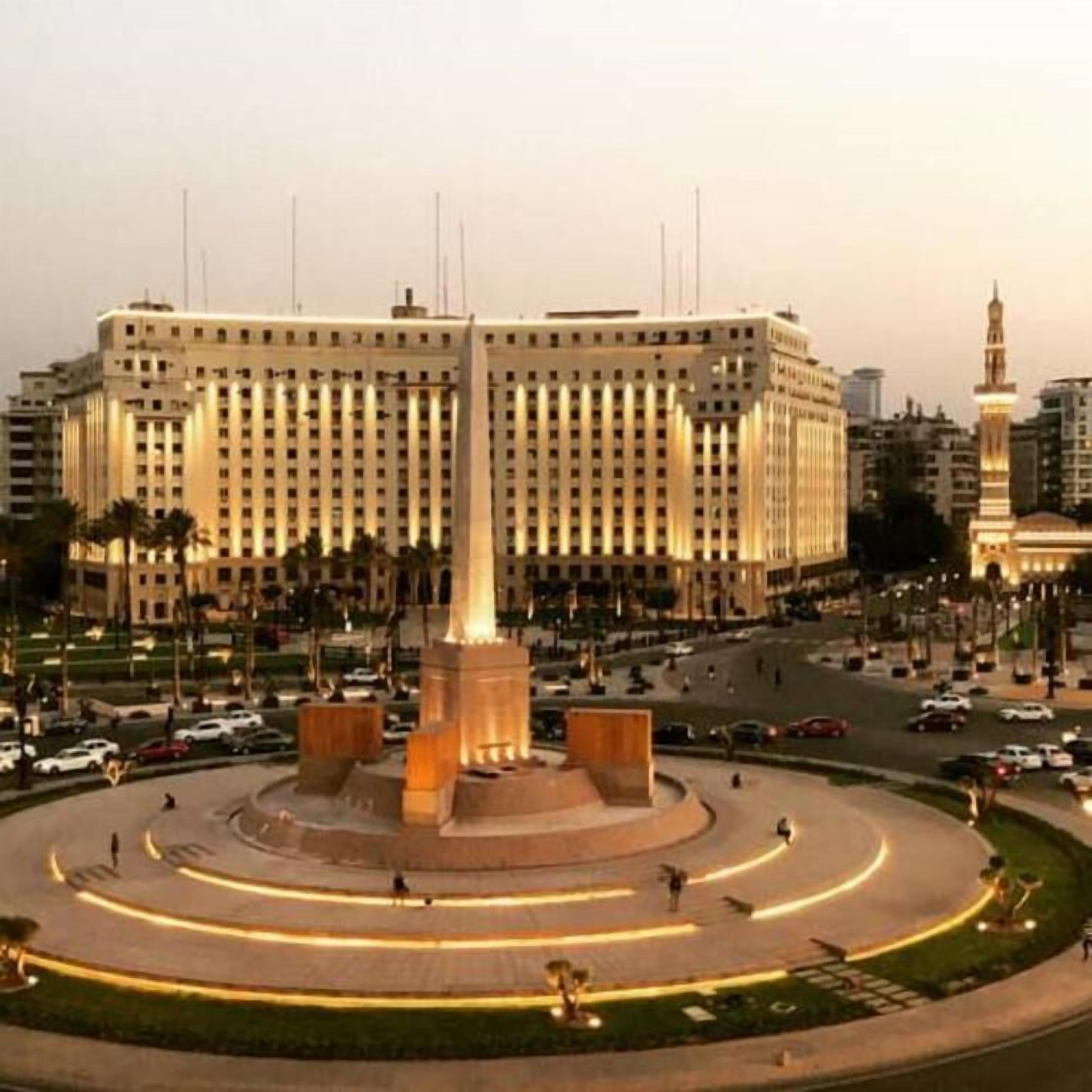 Um edifício grande e com vários andares, com um monumento central cercado por uma praça circular com caminhos iluminados.