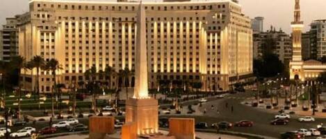Um edifício grande e com vários andares, com um monumento central cercado por uma praça circular com caminhos iluminados.