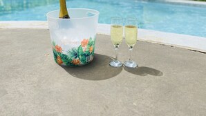 Ambiente à beira da piscina com uma garrafa de champanhe em balde, duas taças de champanhe e uma bola de praia ao fundo.