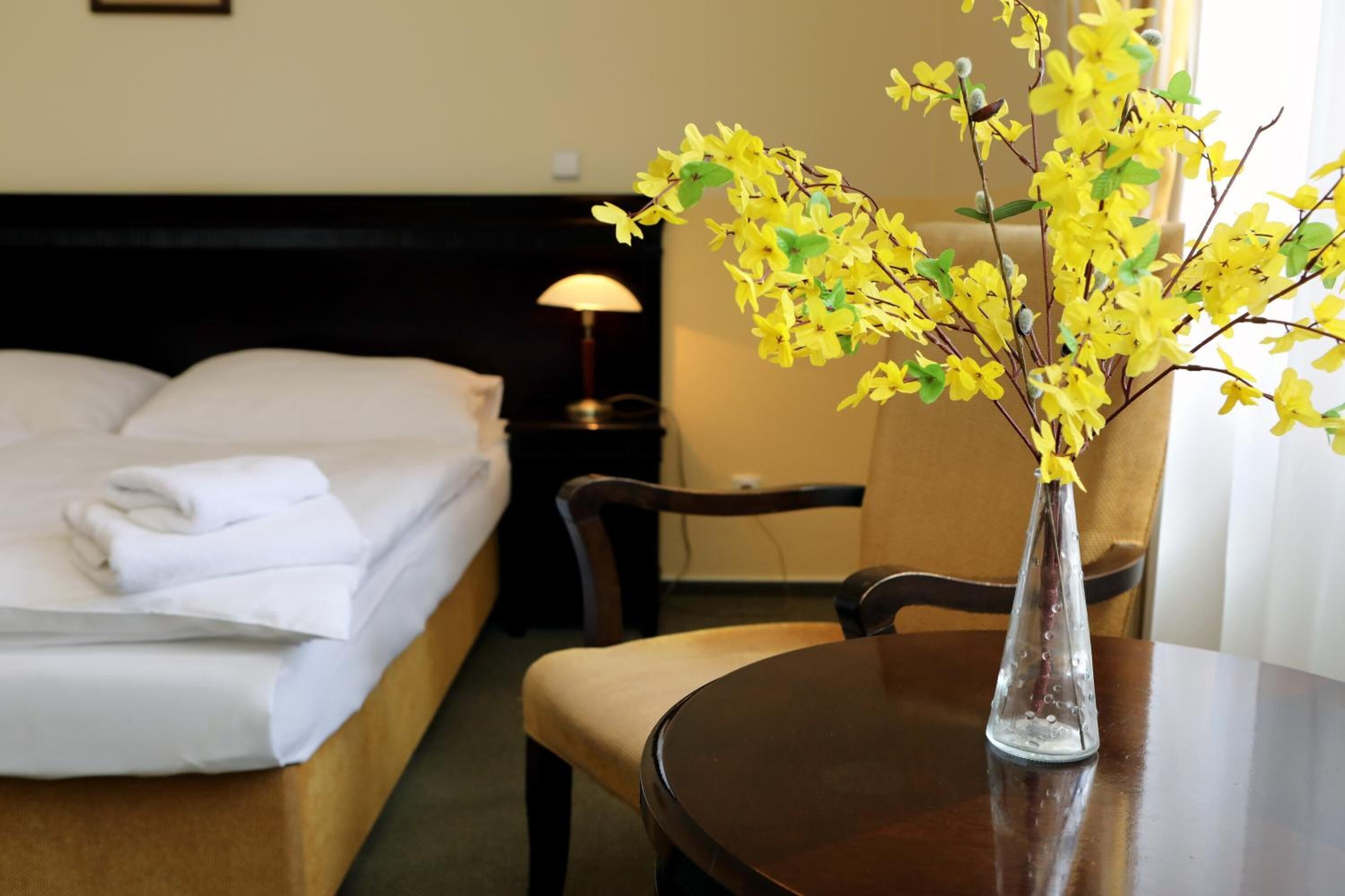 Quarto de hotel com uma cama bem arrumada, um vaso com flores amarelas sobre uma mesa e uma cadeira.