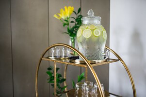 Um frasco de vidro com tampa, cheio de líquido transparente e rodelas de limão, apoiado em um suporte de metal dourado com um tabuleiro contendo vários copos.