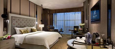 Quarto de hotel com uma cama grande, mesa de cabeceira, cadeira, escrivaninha e vista para a cidade pela janela.