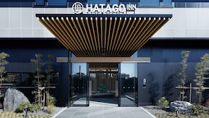 Exterior do edifício HATAGO INN, com design moderno, destacando uma entrada imponente com toldo de ripas de madeira e portas de vidro.