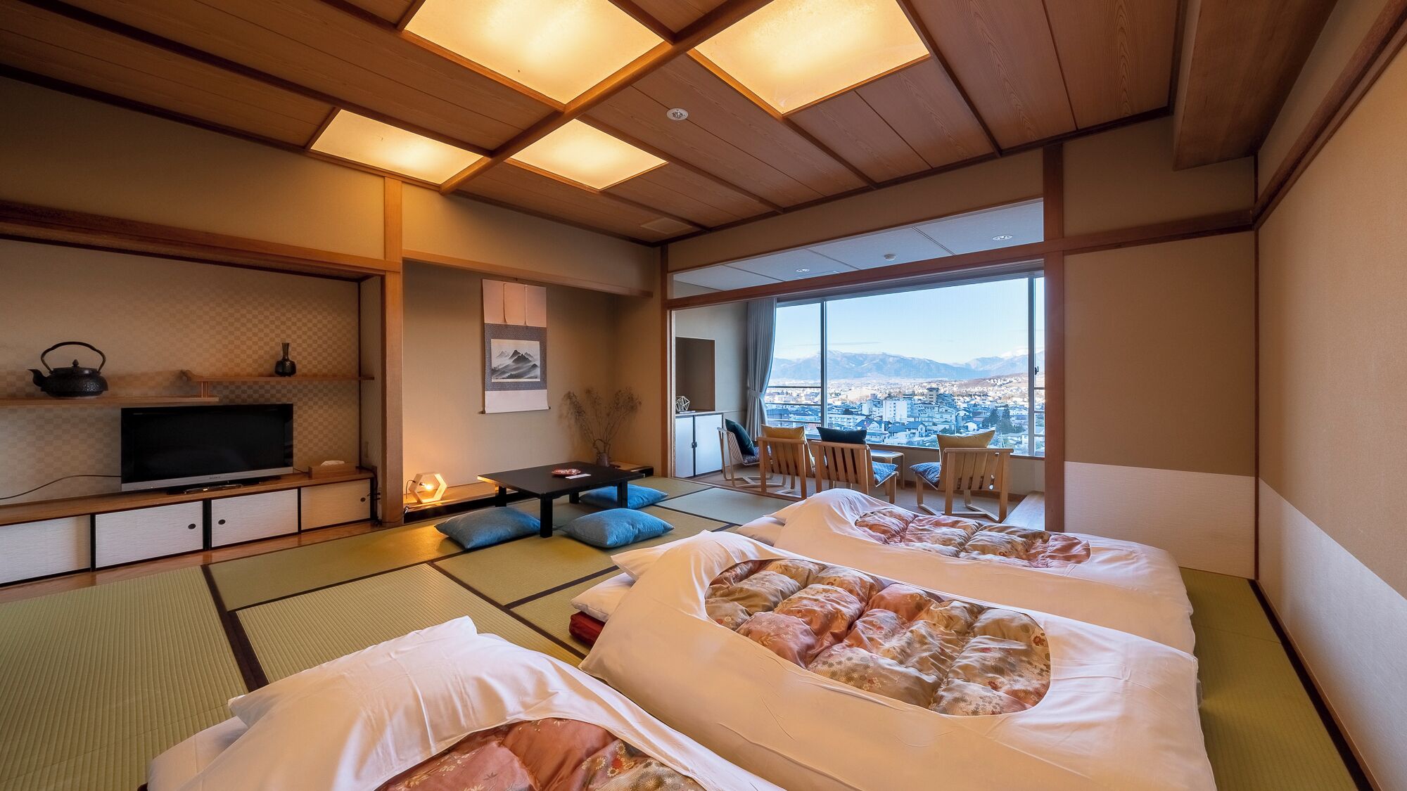Quarto japonês tradicional com tatames, televisão, escrivaninha e vista para a cidade.