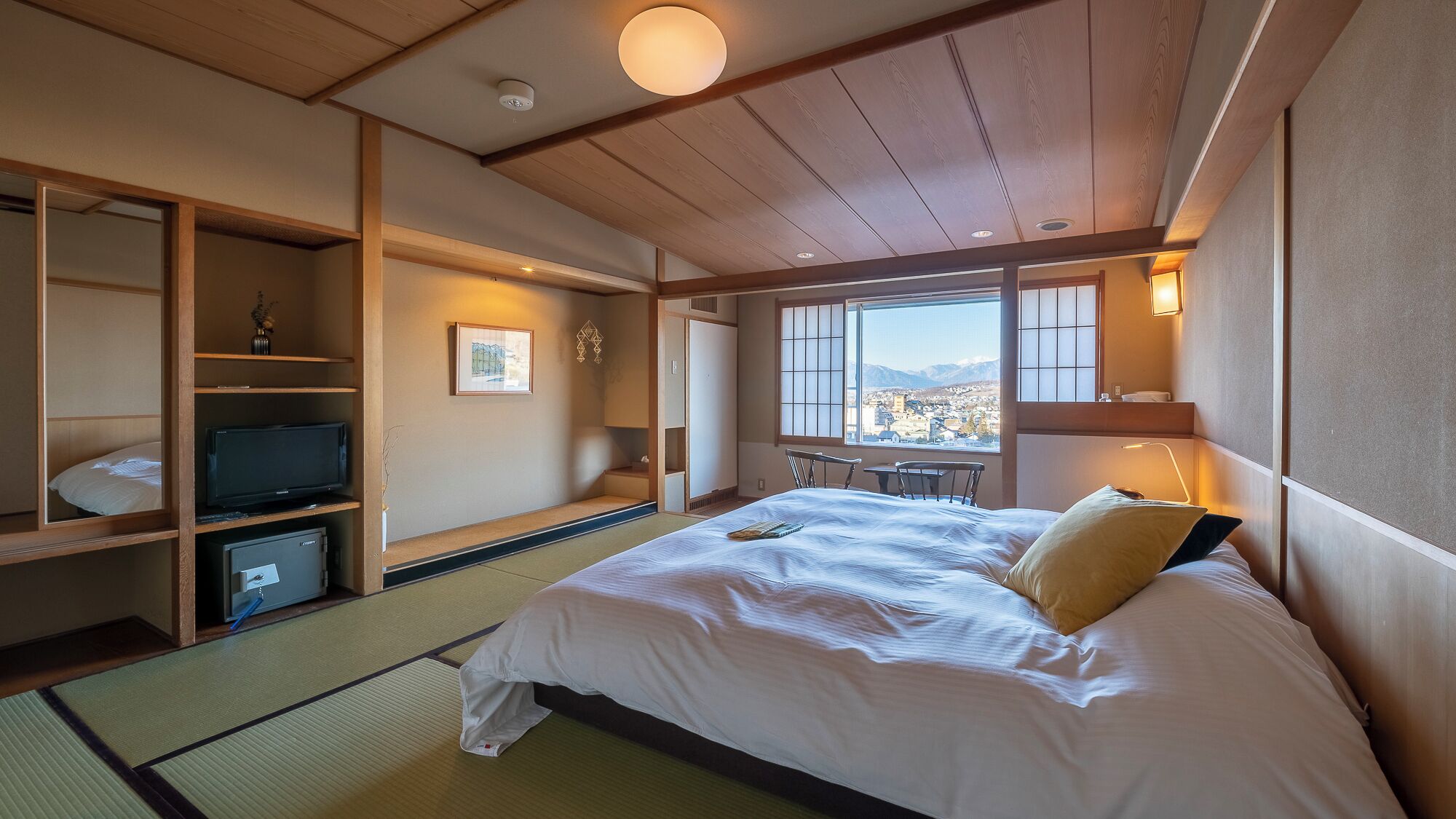 Quarto de hotel tradicional em estilo japonês, com piso de tatame, cama com almofadas, televisão e vista para o horizonte urbano por grandes janelas.