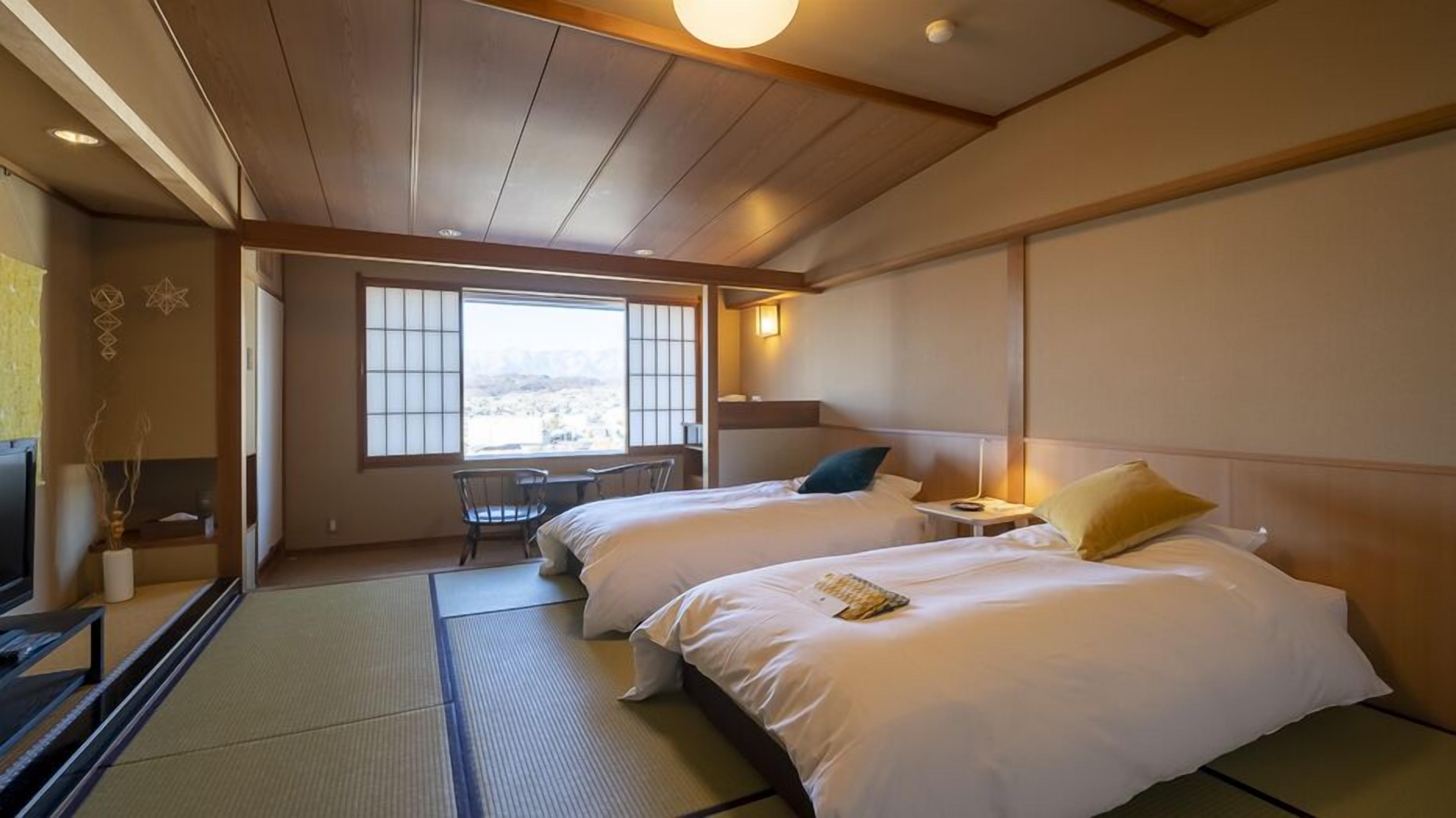 Quarto de hotel em estilo japonês tradicional, com tatames, duas camas, uma escrivaninha e vista para uma paisagem montanhosa.