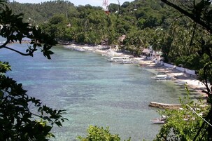Vista costeira com água cristalina, barcos e uma área de praia com edifícios e árvores.