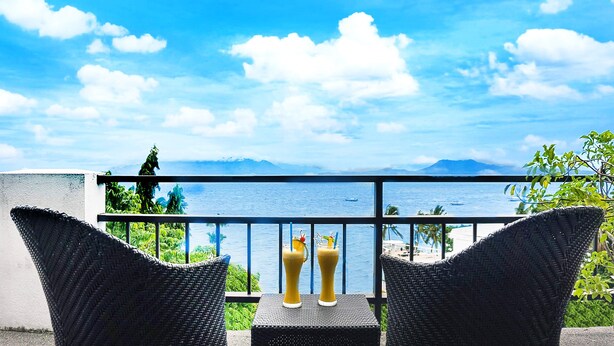 Uma varanda com cadeiras de vime e uma mesa, com vista para o mar e montanhas.