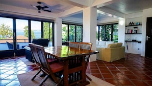 Uma sala de estar ampla com uma mesa de jantar de madeira, assentos confortáveis, grandes janelas com vista para o oceano e um ventilador de teto.