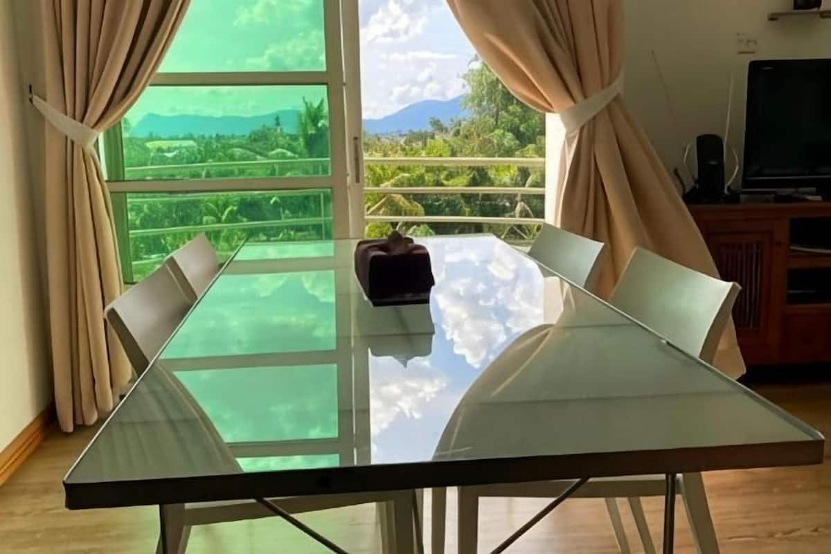 Mesa de jantar com cadeiras, tampo de vidro e vista para o verde pela janela.