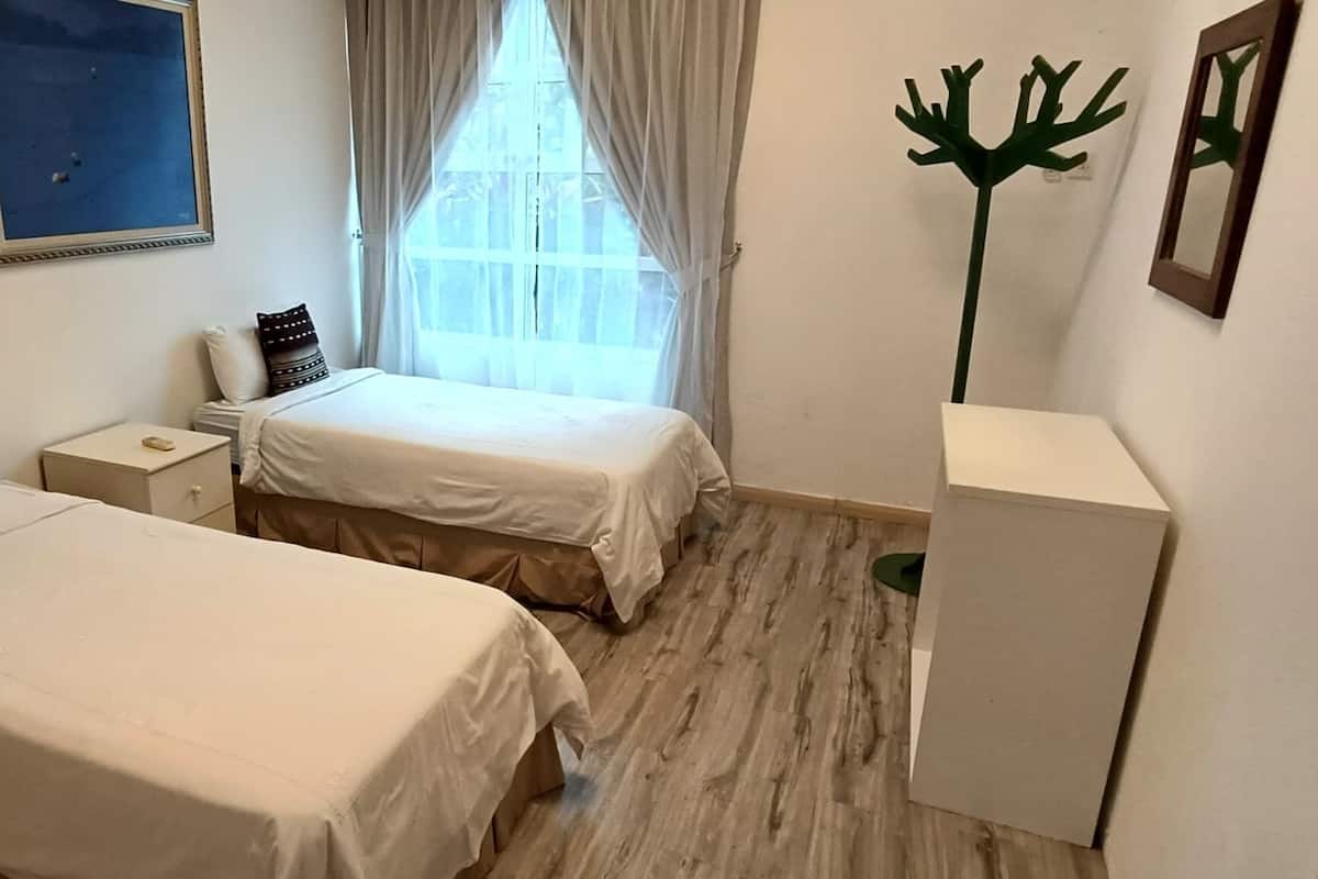 Quarto de hotel com duas camas, uma mesa de cabeceira, uma janela com cortinas, um quadro e um espelho.