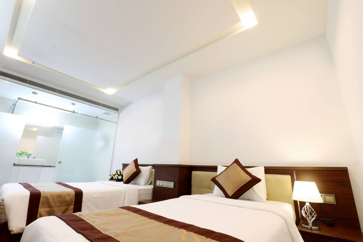 Quarto de hotel com duas camas, mesa de cabeceira com abajur, espelho e janela com vista para o verde.