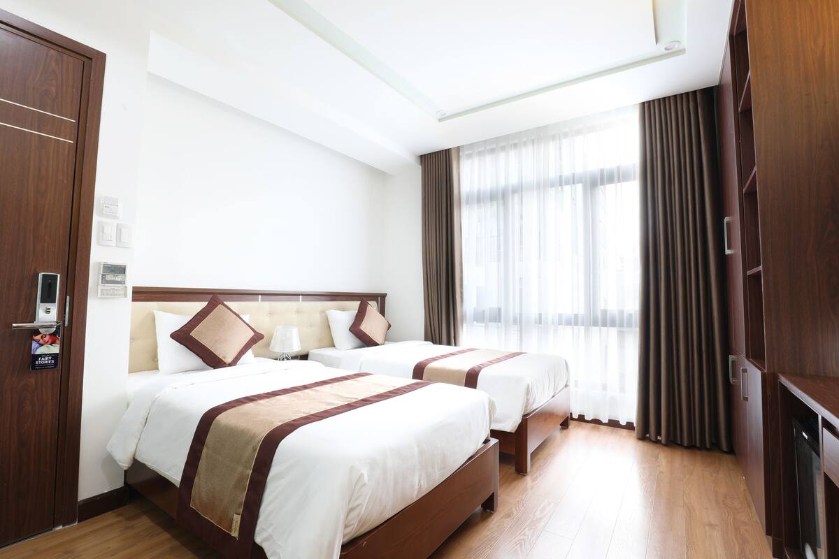 Quarto de hotel com duas camas, um guarda-roupa de madeira, uma escrivaninha e uma janela grande com cortinas.
