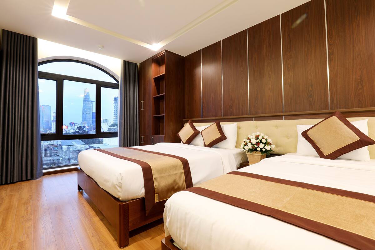 Quarto de hotel com duas camas, painéis de madeira, uma janela grande com vista para a cidade e um arranjo floral na mesa de cabeceira.