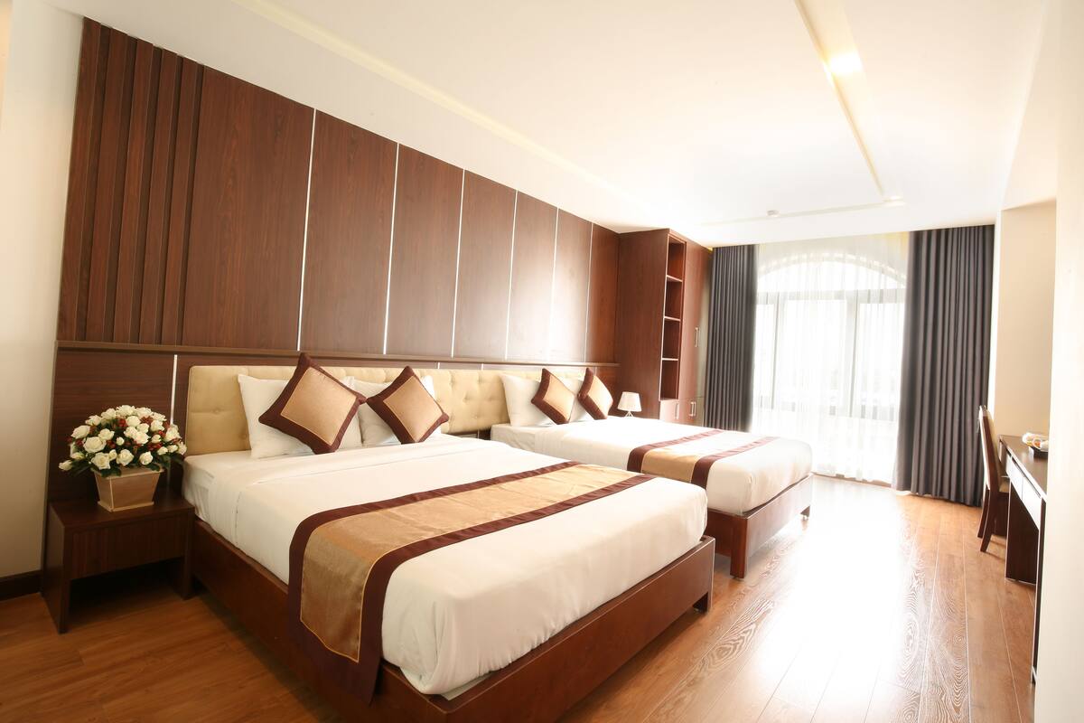 Quarto de hotel com duas camas, painéis de madeira nas paredes, uma mesa de trabalho e uma janela grande com cortinas.