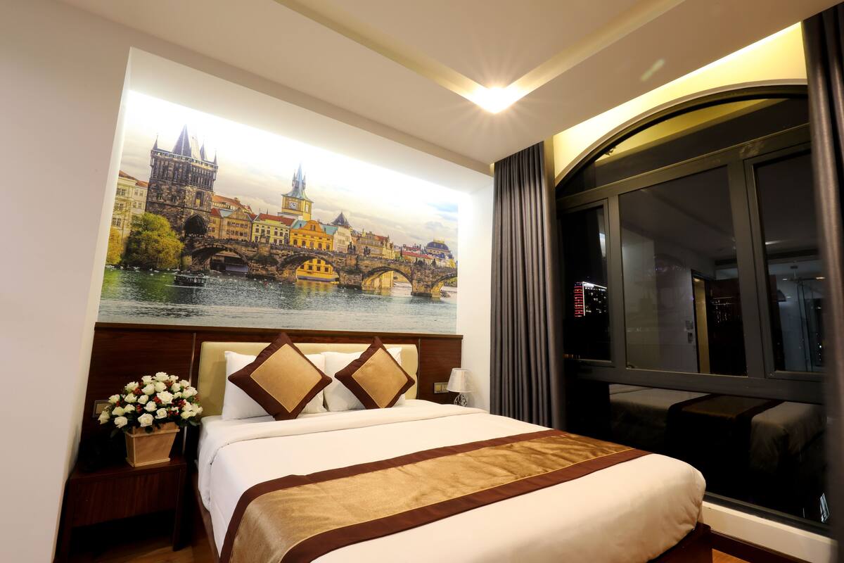 Quarto de hotel com uma cama grande, mesa de cabeceira com um vaso de flores, janela com cortinas e um mural de paisagem urbana na parede.