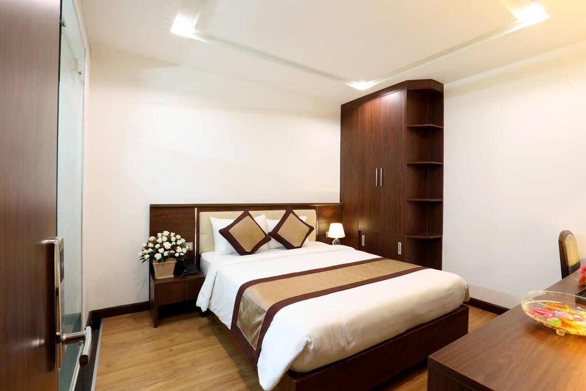 Quarto de hotel com uma cama grande, guarda-roupa de madeira, mesa de cabeceira com abajur e uma pequena escrivaninha com objeto decorativo.