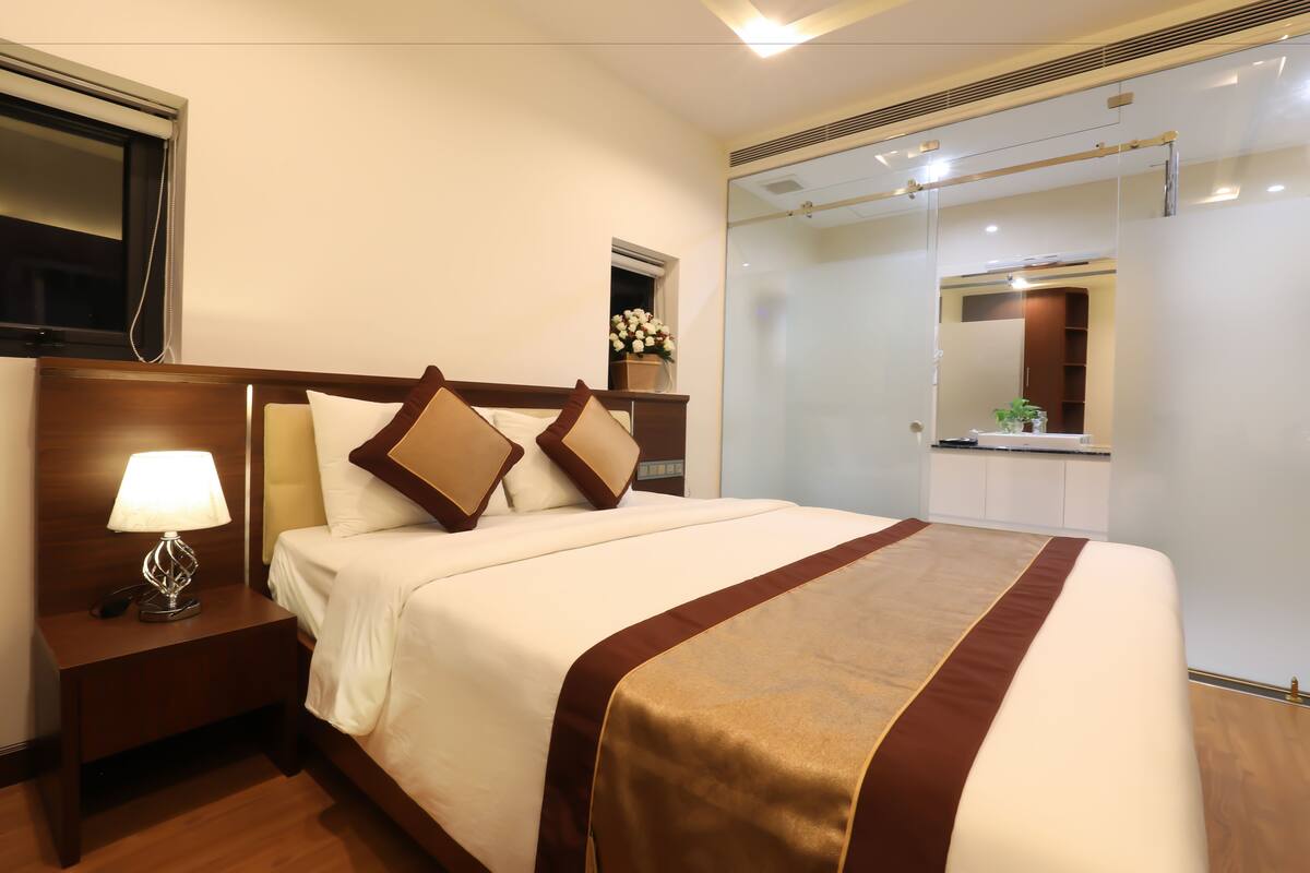 Quarto de hotel com uma cama grande, mesa de cabeceira com abajur, espelho e uma pequena janela com vista para o exterior.