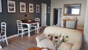Uma área de refeições com uma mesa e cadeiras brancas, fotografias em preto e branco nas paredes, um espelho sobre a mesa e um sofá com um vaso de flores ao primeiro plano.