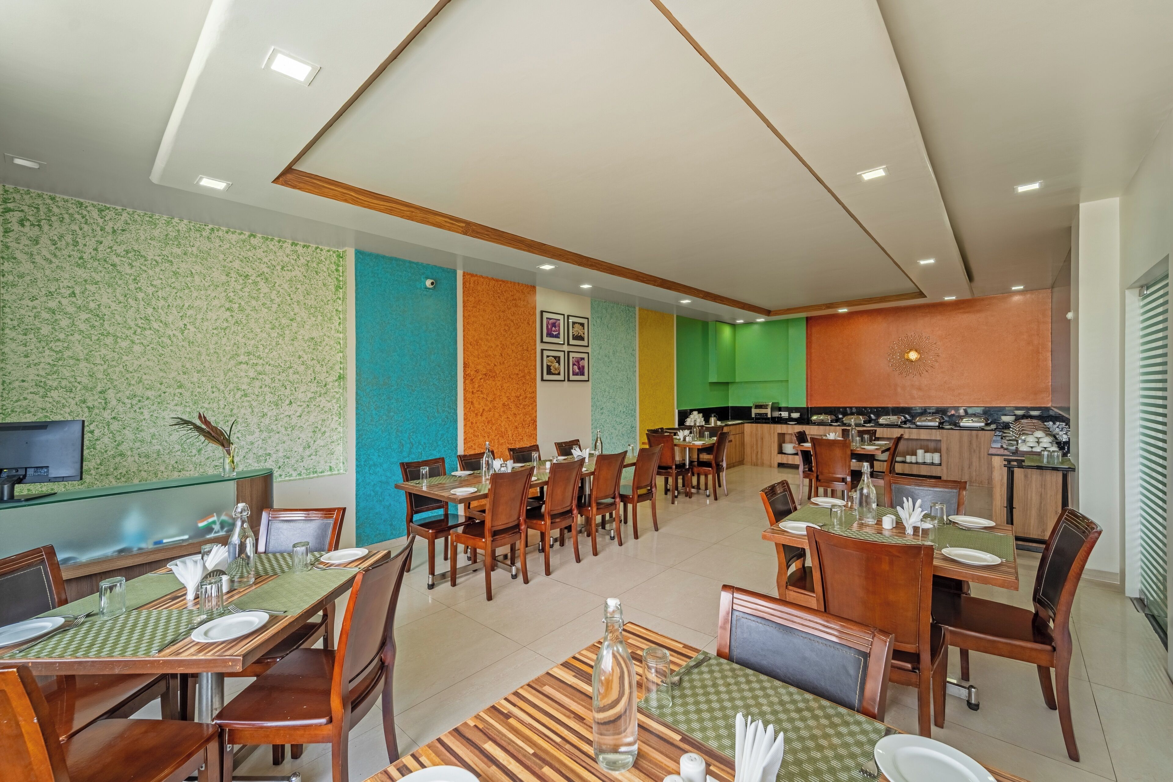Uma sala de jantar com várias mesas e cadeiras, uma cozinha ao fundo e uma televisão à esquerda.