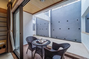 Uma varanda com cadeiras de vime, uma mesa com um tabuleiro de chávenas e um livro, e vista para o exterior do edifício.