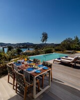 Uma área de refeições à beira da piscina, com uma mesa posta para dois, com vista para a piscina e uma paisagem cénica de colinas e um lago.