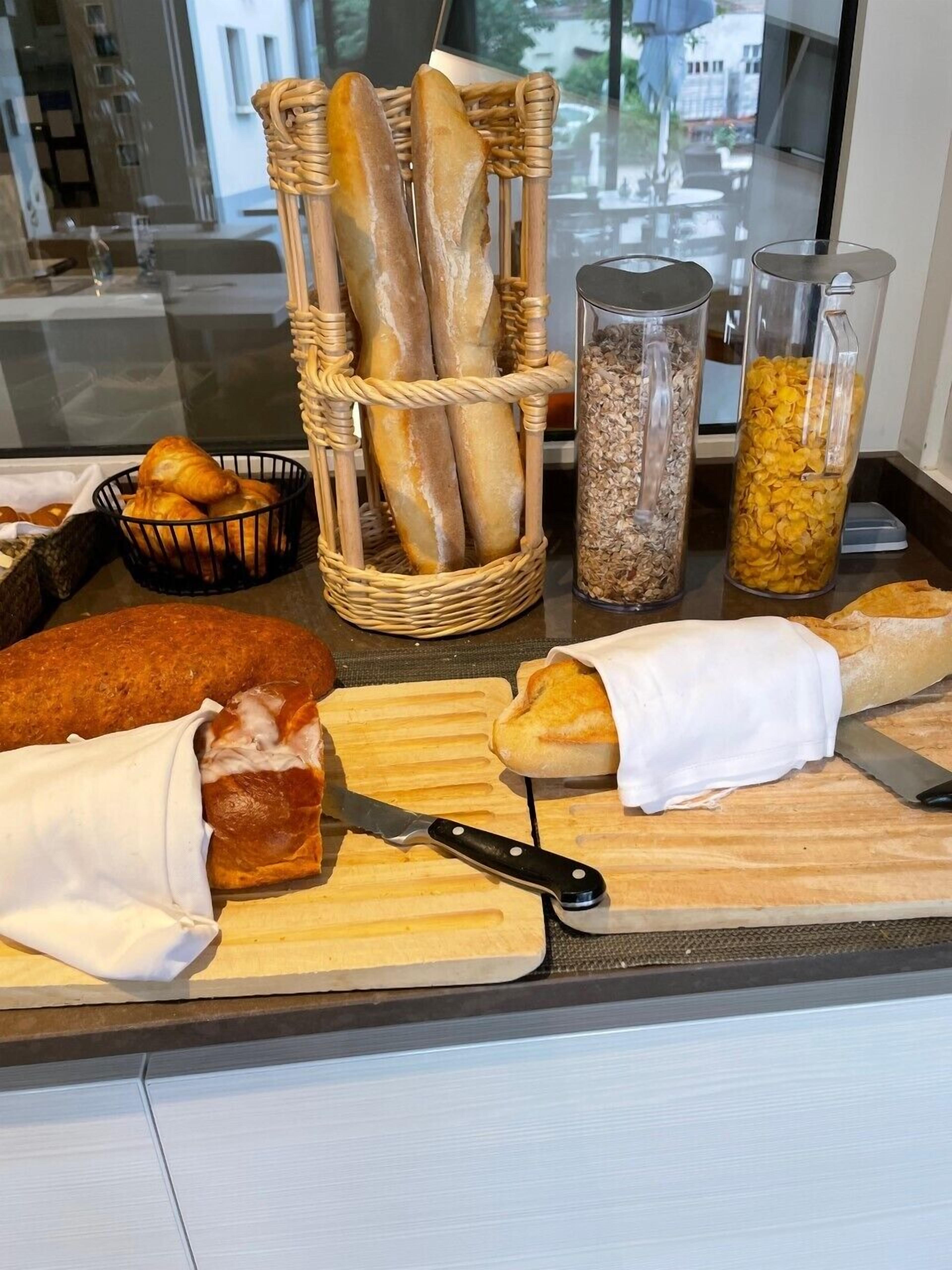 Uma bancada de cozinha com vários pães, um cesto de croissants, um pote de aveia e um pote de sementes de girassol.