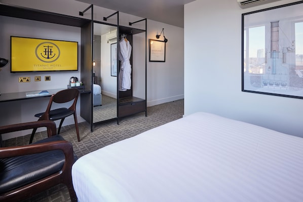 Quarto de hotel com uma cama, uma secretária com um computador, uma cadeira, um roupão pendurado em um cabide e um quadro com a imagem de um edifício.
