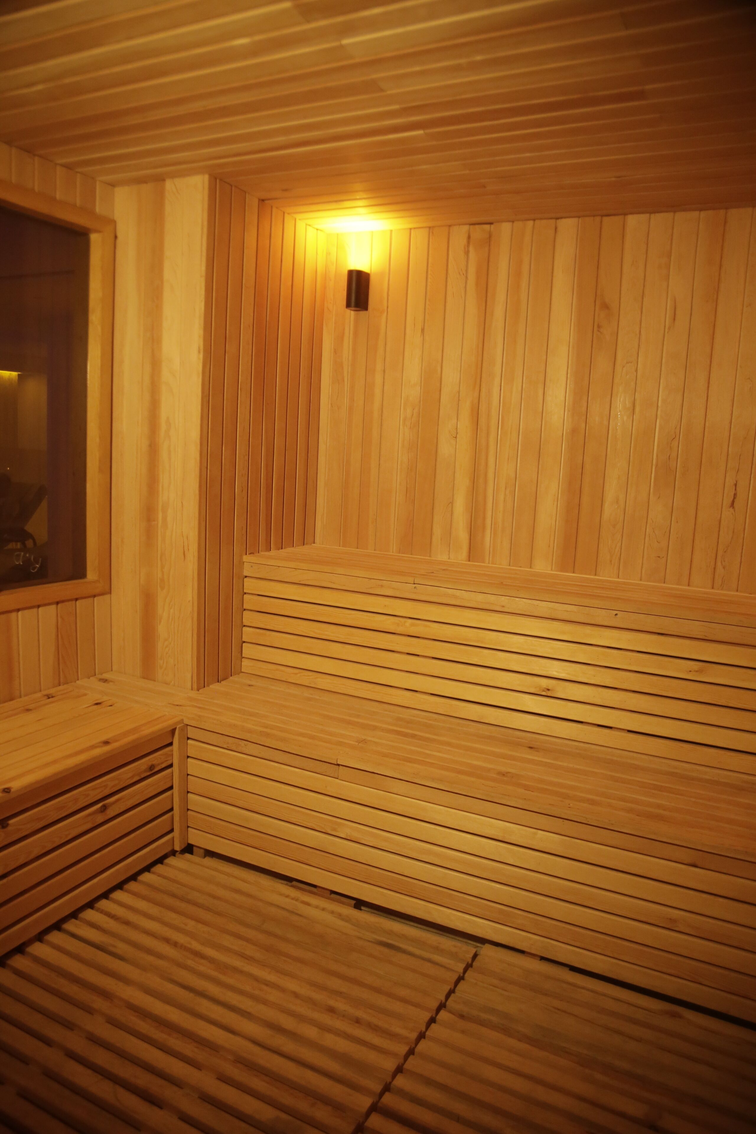 Uma sauna de madeira com bancos e assento de banco, uma janela e um aparelho de luz fixado na parede.