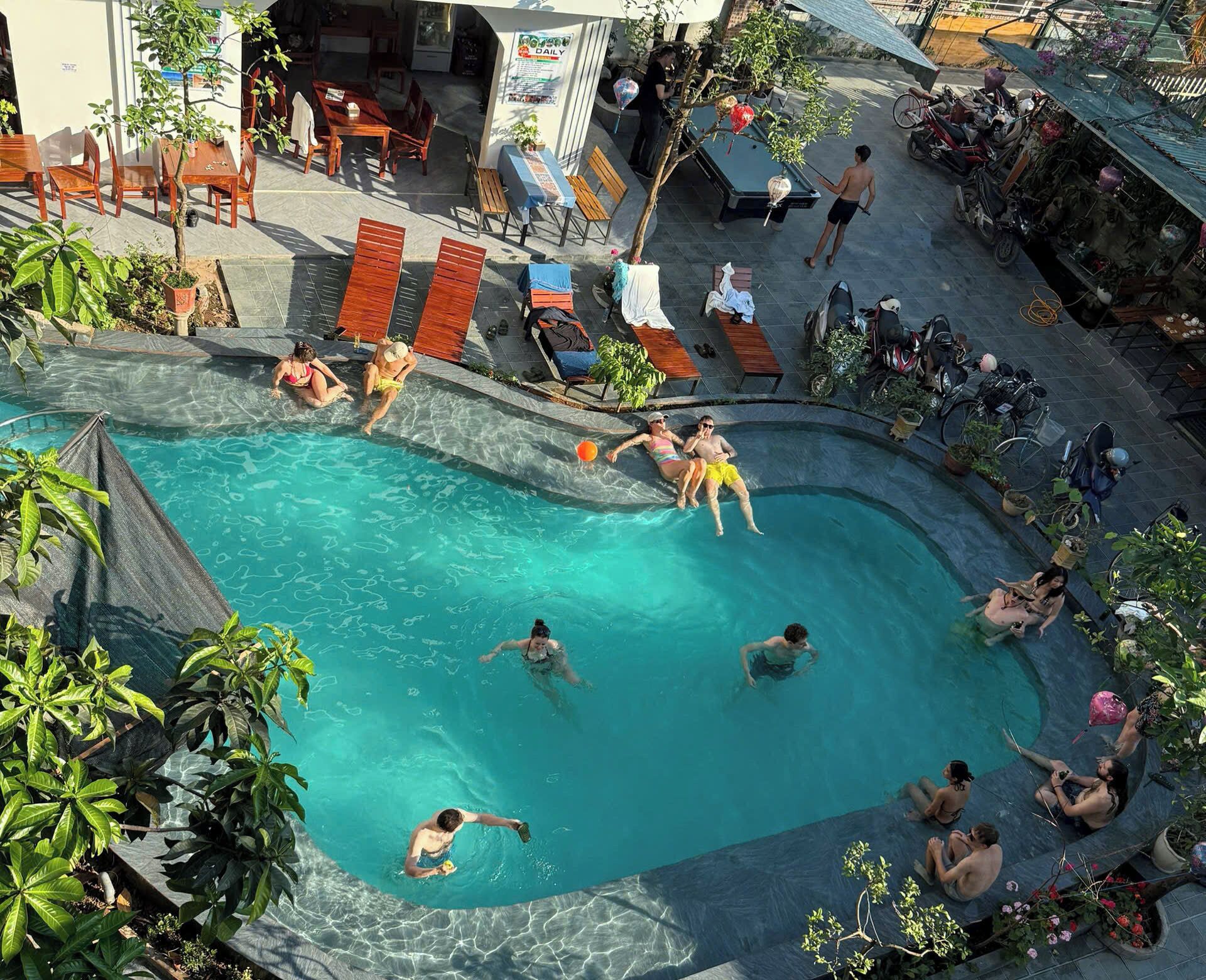 Piscina com pessoas nadando e relaxando. Cadeiras e mesas estão dispostas na área externa da piscina.