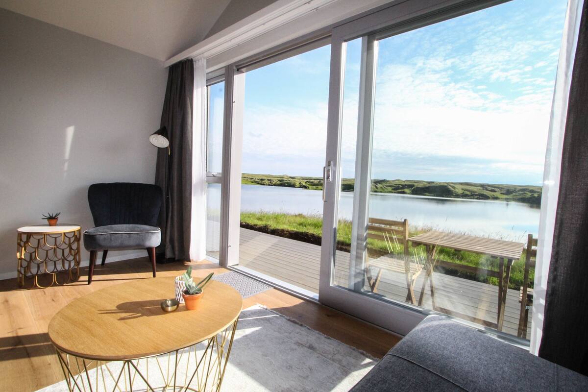 Quarto moderno com uma grande porta de correr de vidro que dá para um deck com vista para um lago e colinas gramadas.