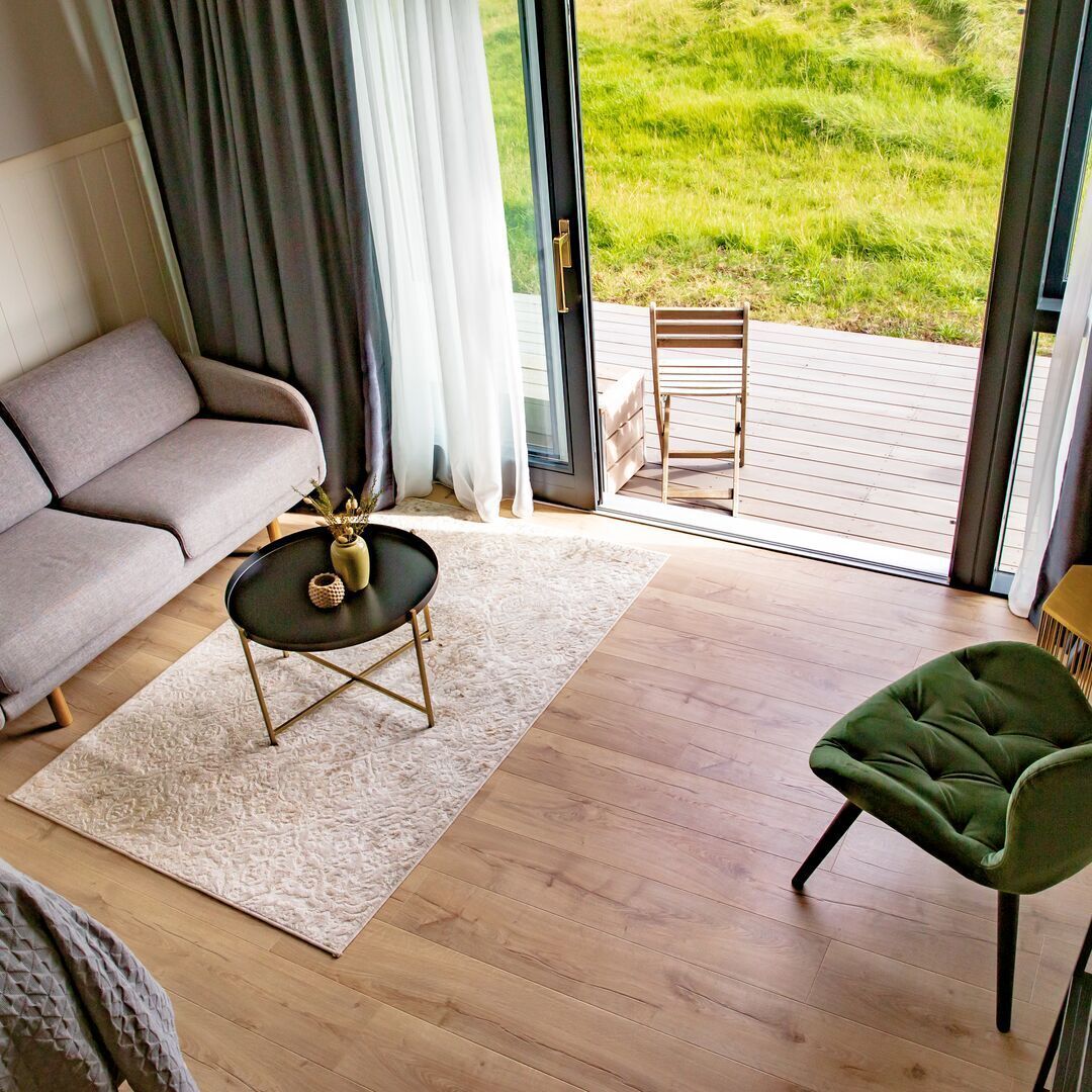 Uma sala de estar moderna com um sofá, uma poltrona verde, uma mesinha com objetos decorativos, um tapete branco e uma porta de vidro deslizante que dá para um deck externo.