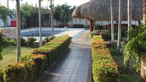 Uma área de piscina com um caminho pavimentado, palmeiras e uma estrutura com telhado de palha.