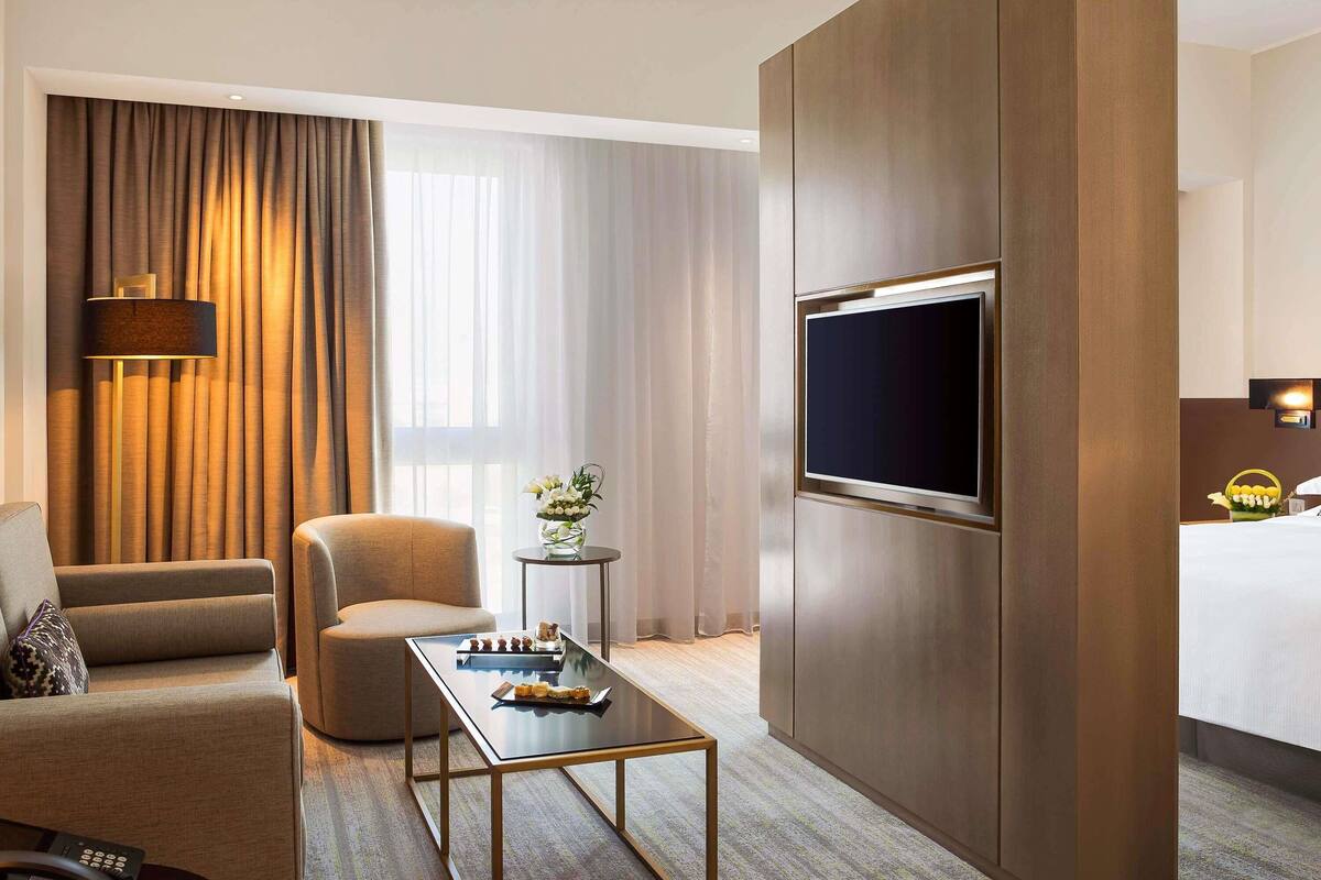 Quarto de hotel com sofá, poltrona, mesa de centro, televisão e uma pequena mesa lateral com flores.