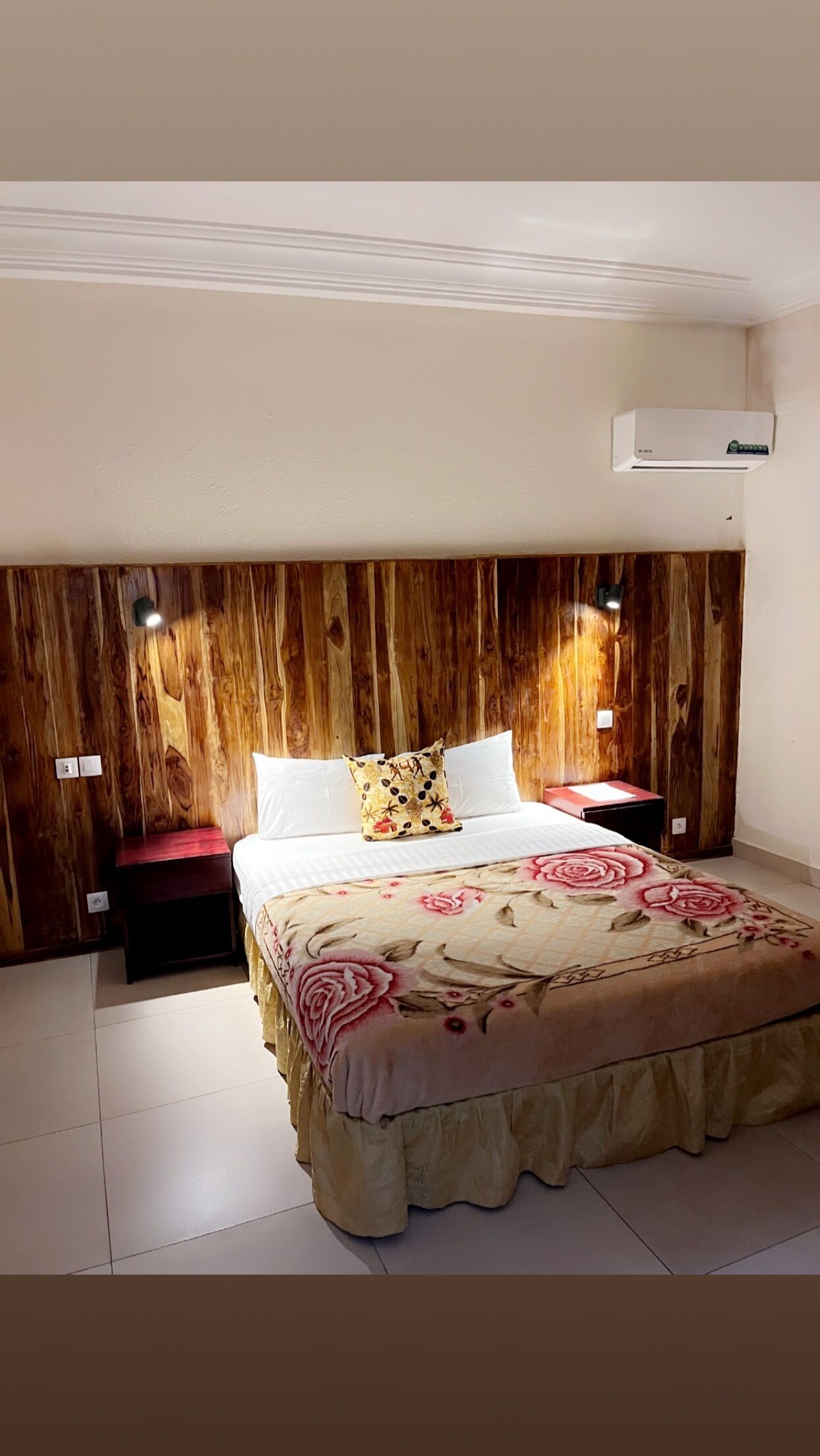 Quarto com cabeceira de madeira, cama com roupa de cama floral, duas mesas de cabeceira e um aparelho de ar condicionado na parede.