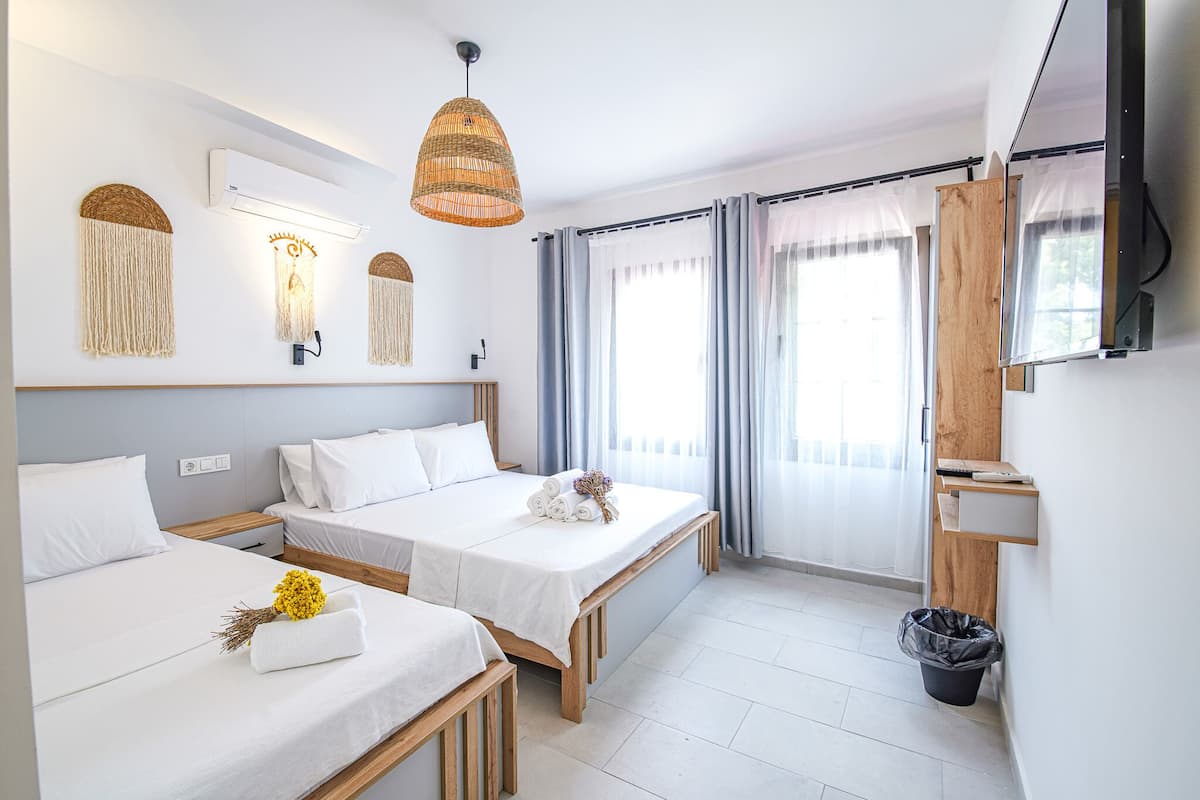 Quarto de hotel com duas camas de solteiro, um banco de madeira, uma lixeira, uma televisão presa à parede e uma janela grande com cortinas.