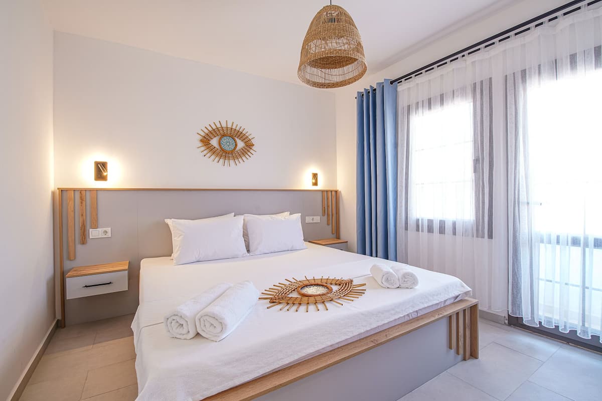 Quarto de hotel moderno com uma cama grande, mesa de cabeceira, luminária na parede e janela com cortinas.