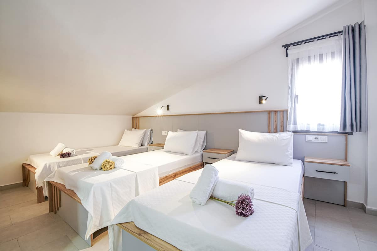 Quarto de hotel com três camas dispostas em linha, cada uma com roupa de cama e almofadas brancas. Há uma janela com cortinas no lado direito.