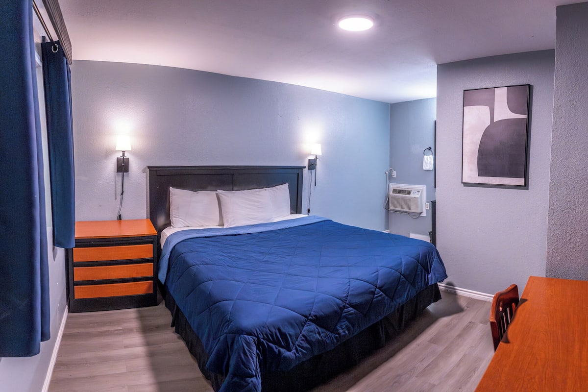 Quarto de hotel com cama, mesa de cabeceira, luminária, ar condicionado e escrivaninha com cadeira.
