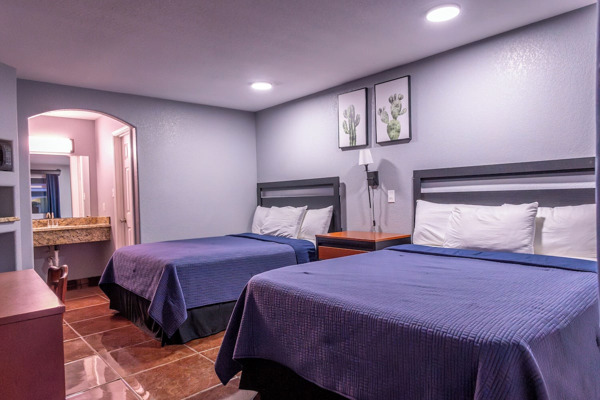 Quarto de hotel com duas camas, uma escrivaninha, uma cadeira, toucador com espelho e duas fotos emolduradas na parede.