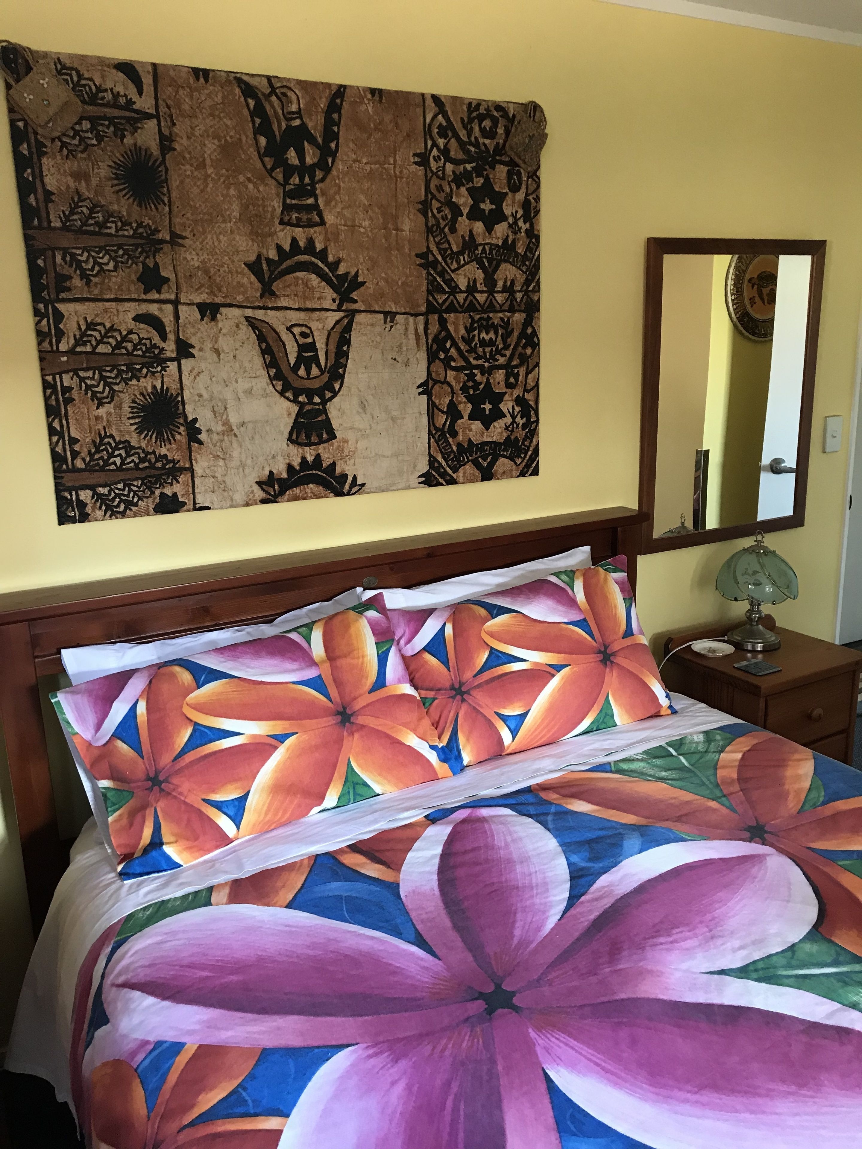 Uma cama com um cobre-leito floral colorido, uma tapeçaria de parede com desenhos detalhados e um espelho sobre uma mesa de cabeceira de madeira.
