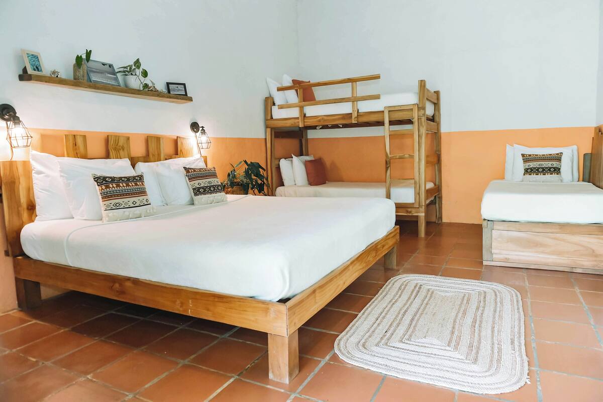 Quarto com beliche de madeira, uma cama de solteiro e uma escrivaninha pequena. Há prateleiras sobre as camas com objetos decorativos e um vaso de planta.