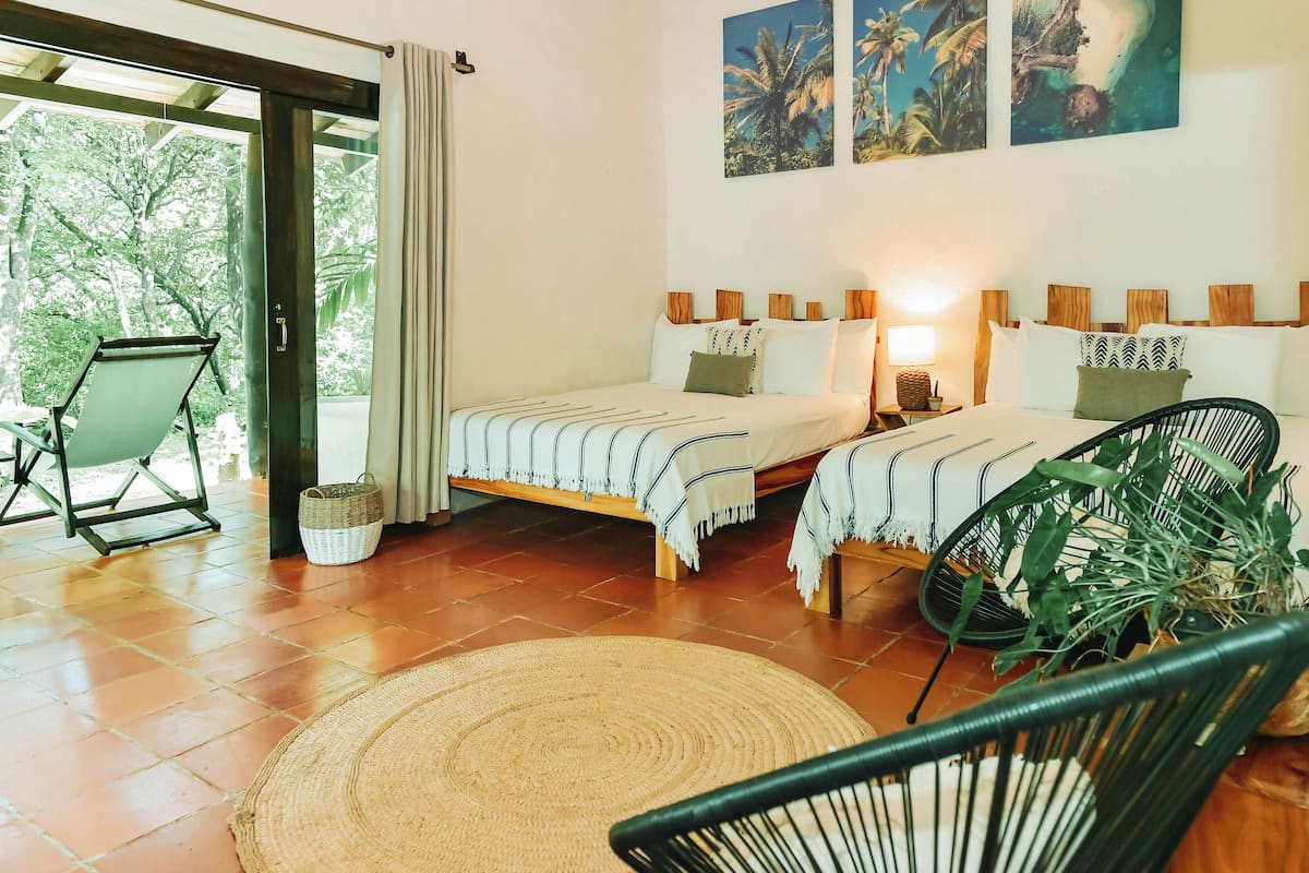 Quarto com duas camas, uma cadeira verde, um tapete tecido e vista para árvores através de uma porta de vidro.