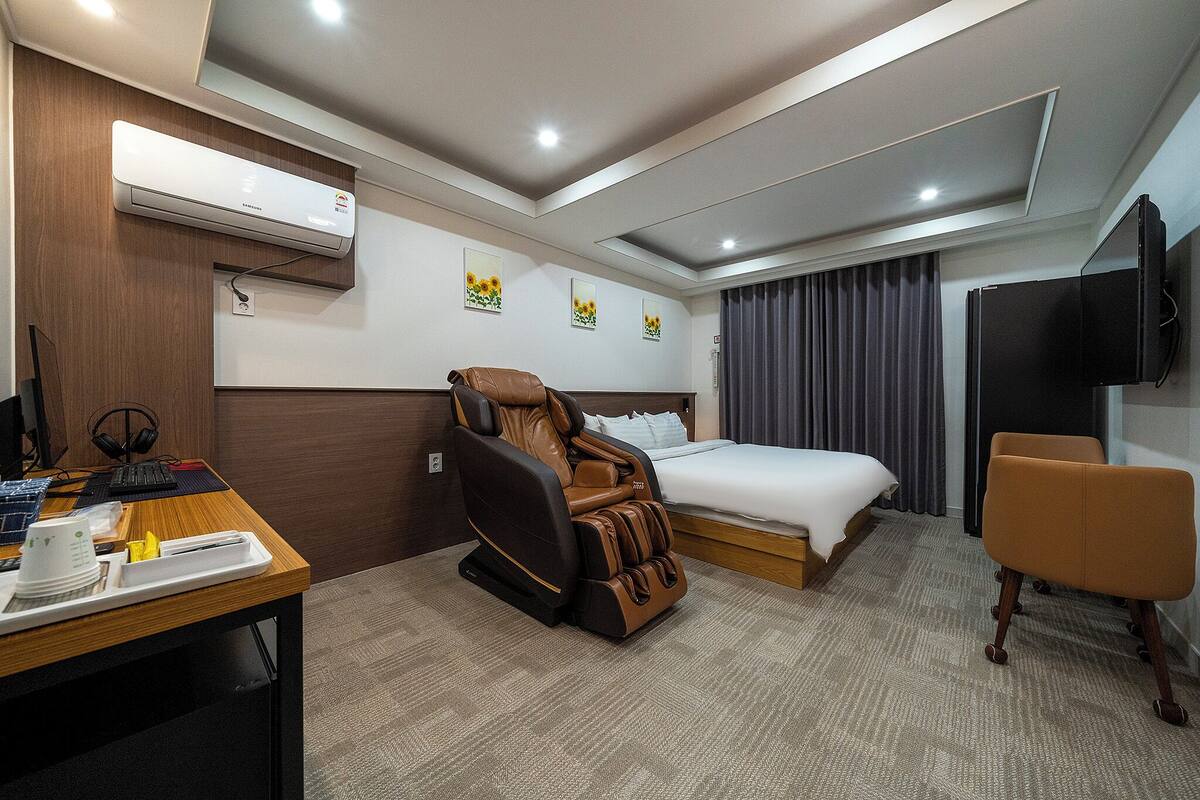 Quarto de hotel com cadeira de massagem, cama, escrivaninha e computador, e televisão.