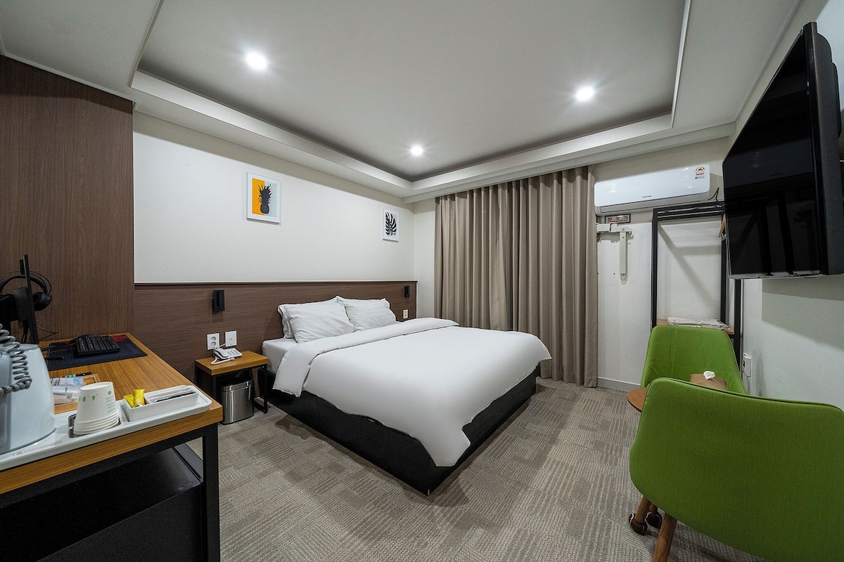 Quarto de hotel com cama, escrivaninha com telefone, lixeira, cadeira verde e ar condicionado.