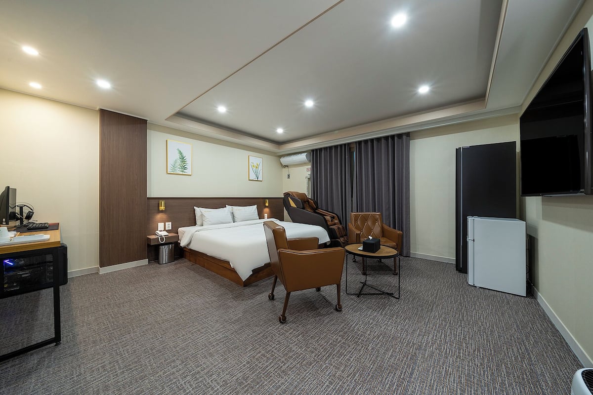 Quarto de hotel com cama, escrivaninha com computador, cadeira, mesa pequena, televisão e refrigerador.