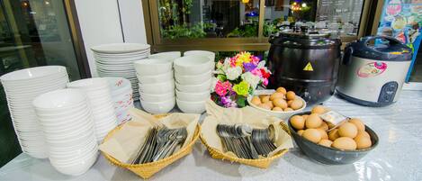 Uma mesa com pilhas de pratos brancos, tigelas e cestos de utensílios, além de uma cafeteira e uma tigela de ovos.