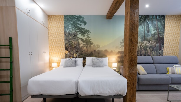 Quarto com uma cama de casal, um sofá cinza e um mural de paisagem tropical.