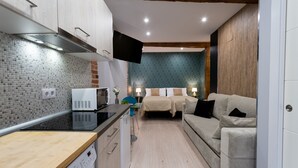Cozinha compacta com micro-ondas, máquina de lavar louça e bancada de madeira. Uma cama com cobertor e travesseiros bege é visível ao fundo.
