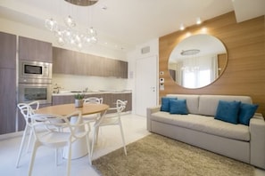 Uma cozinha moderna com eletrodomésticos embutidos, uma área de jantar com mesa e cadeiras redondas, e uma sala de estar com sofá e espelho redondo.